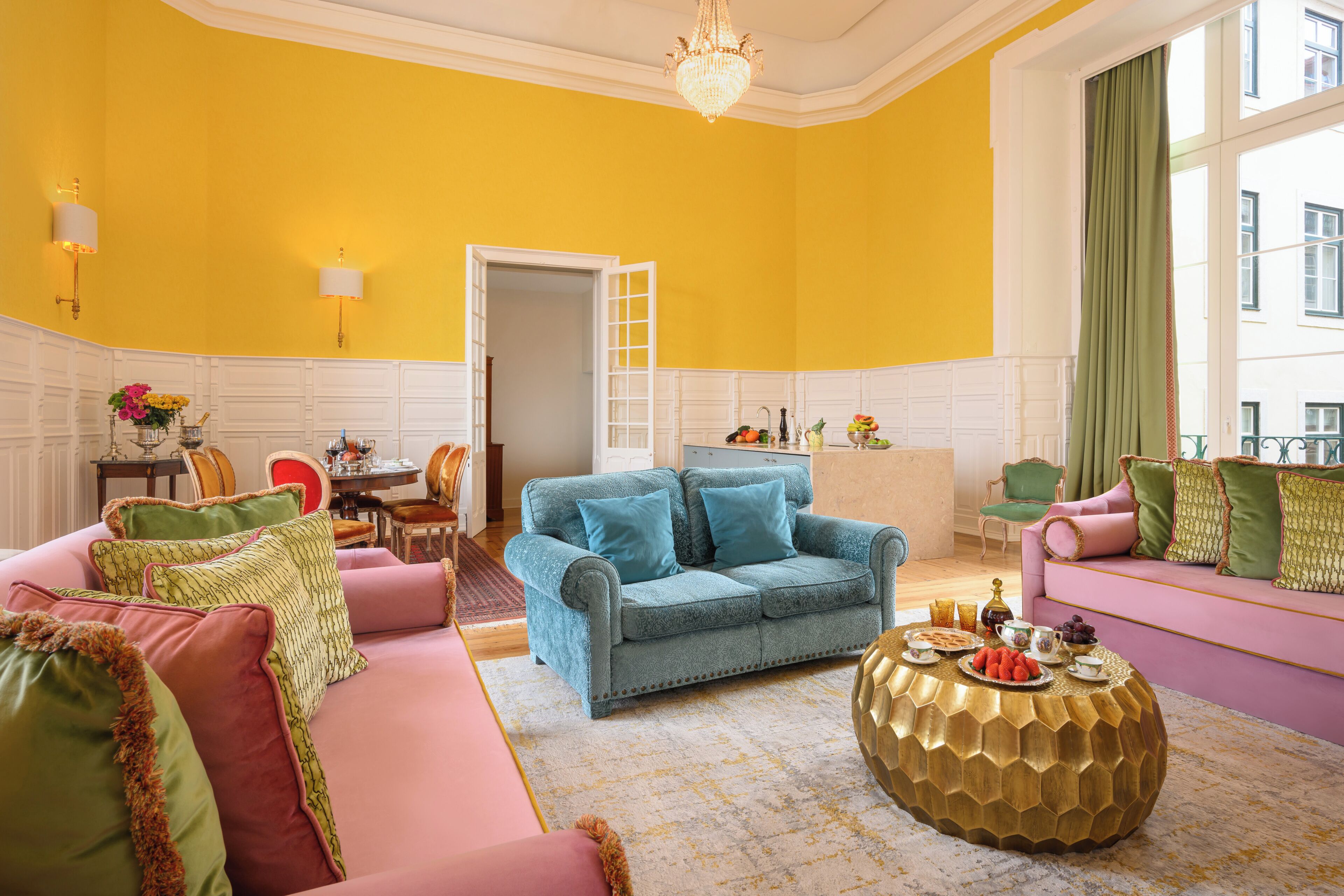 Um quarto com um sofá azul, um sofá rosa, uma mesa de centro dourada e uma parede amarela.