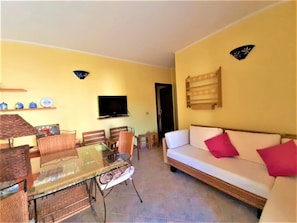 Uma sala de estar com uma mesa de jantar de vidro, cadeiras de madeira, um sofá branco com almofadas vermelhas, uma TV de tela plana na parede e uma pequena estante com objetos decorativos.