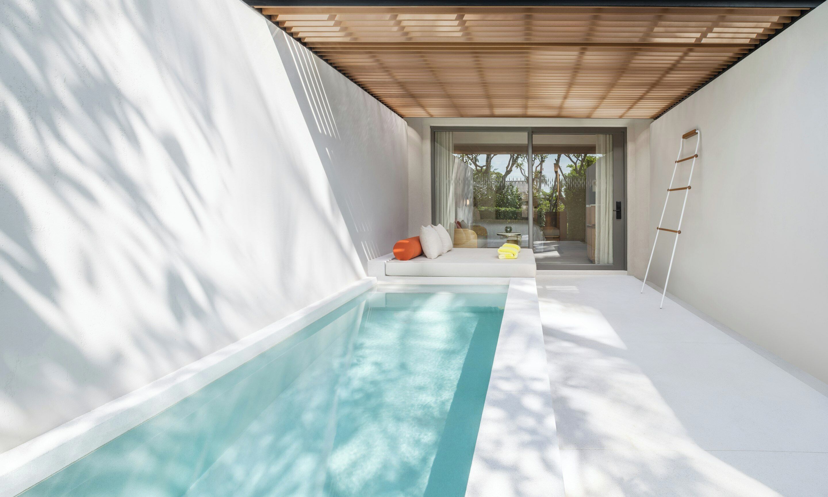 Uma área moderna com piscina ao ar livre, com um pergolado de madeira, espaço de estar com almofadas e porta de vidro que dá para um jardim.