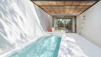 Uma área moderna com piscina ao ar livre, com um pergolado de madeira, espaço de estar com almofadas e porta de vidro que dá para um jardim.