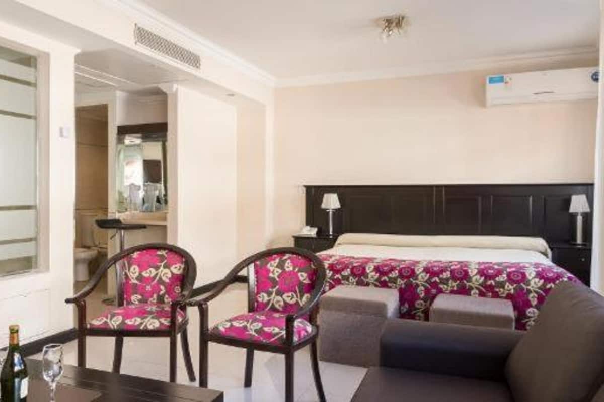 Quarto de hotel com cama, duas cadeiras, sofá, uma mesinha com garrafa e copos, e um espelho.