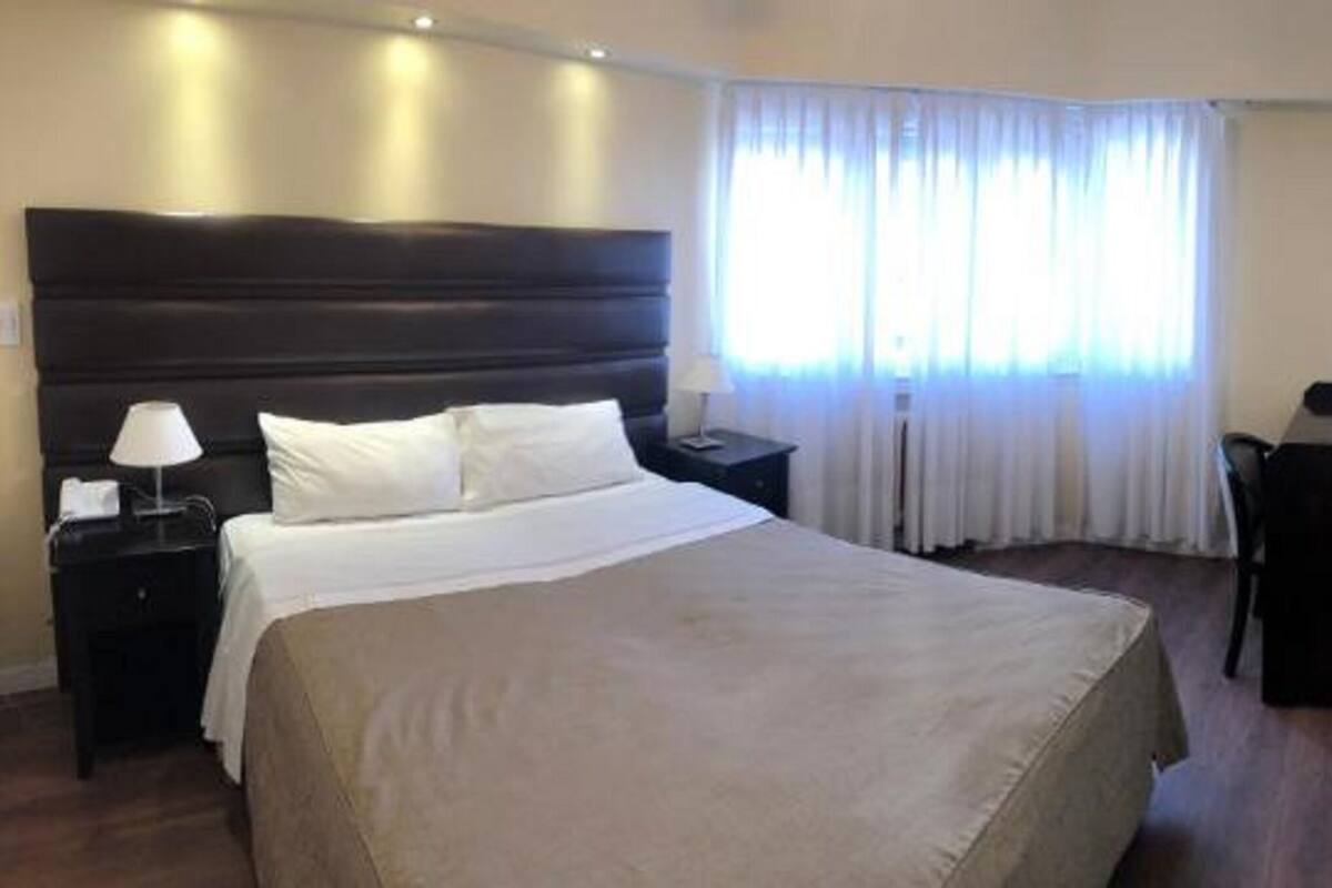 Quarto de hotel com uma cama grande, uma mesa de cabeceira com abajur, uma cadeira e uma janela com cortinas.