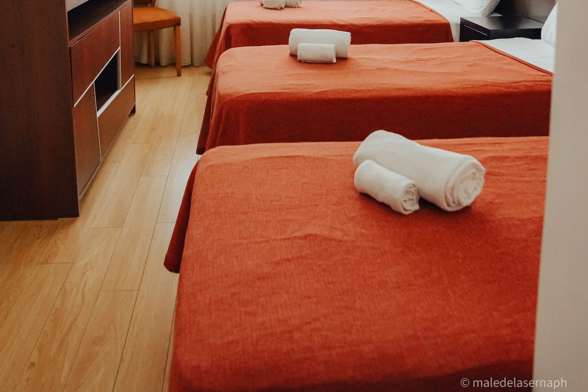 Quarto de hotel com duas camas, uma escrivaninha, uma cadeira, ar condicionado e uma janela com cortinas.