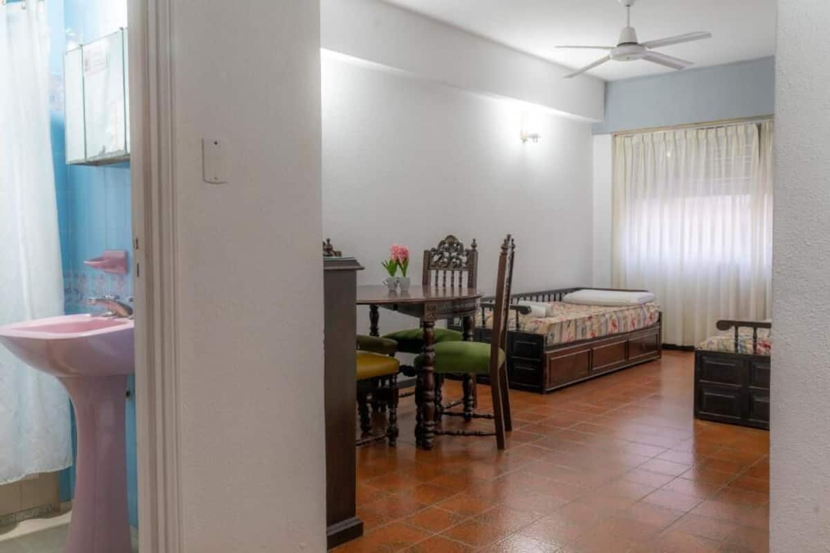 Quarto com cama, escrivaninha, cadeira, toucador com pia e janela com cortinas.