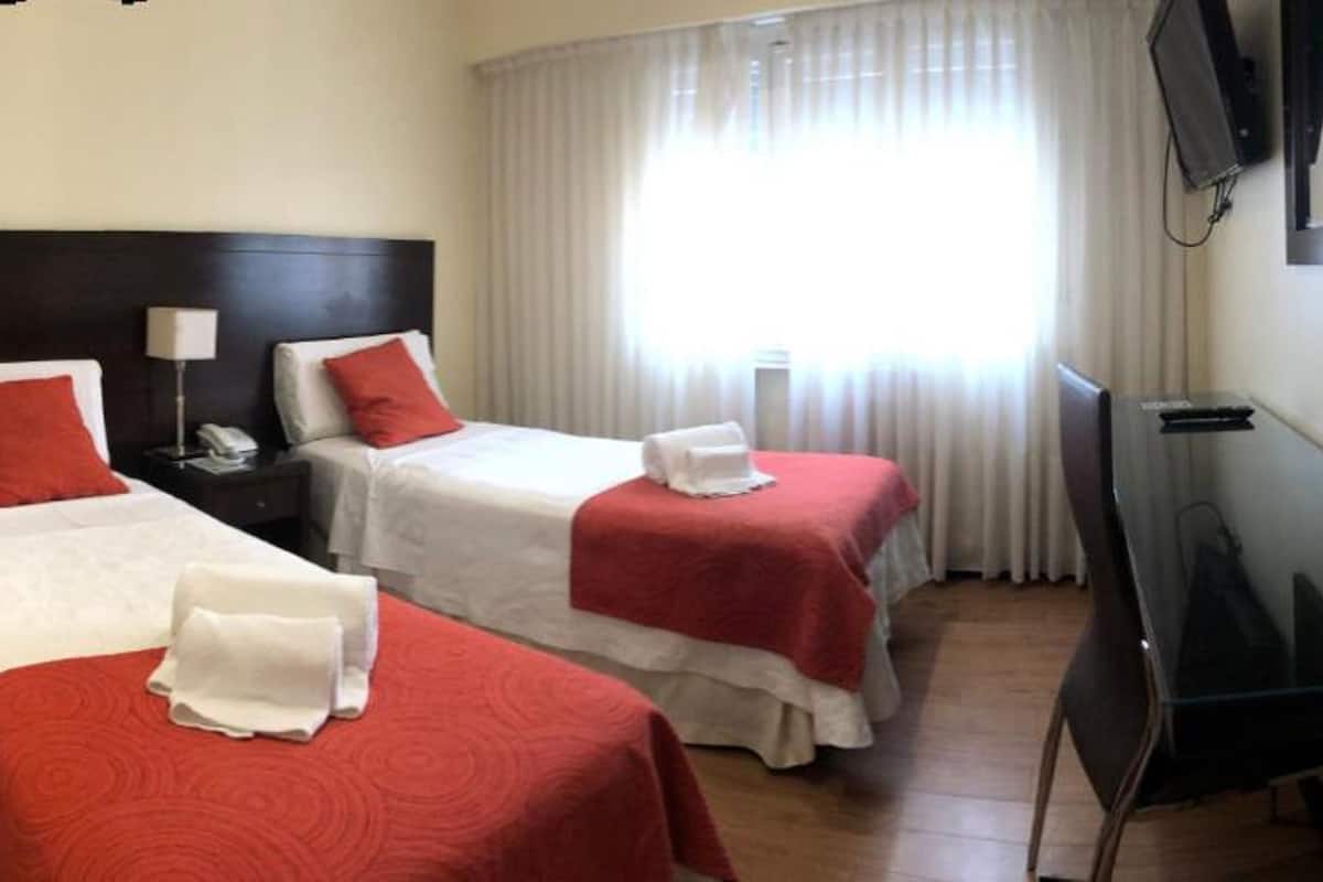 Quarto de hotel com duas camas, cada uma com uma colcha vermelha e branca, uma mesa de cabeceira com abajur e uma televisão na parede.