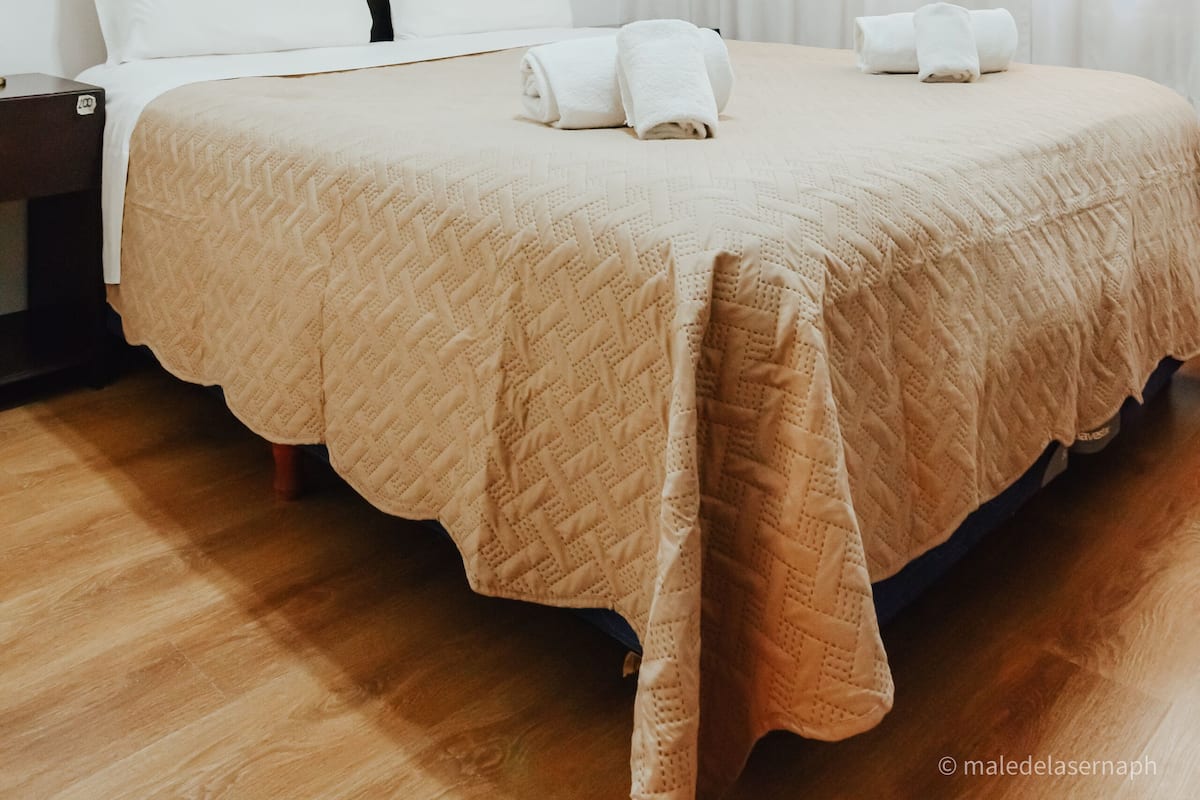 Cama bem arrumada com colcha bege, cabeceira de madeira e cortinas brancas.