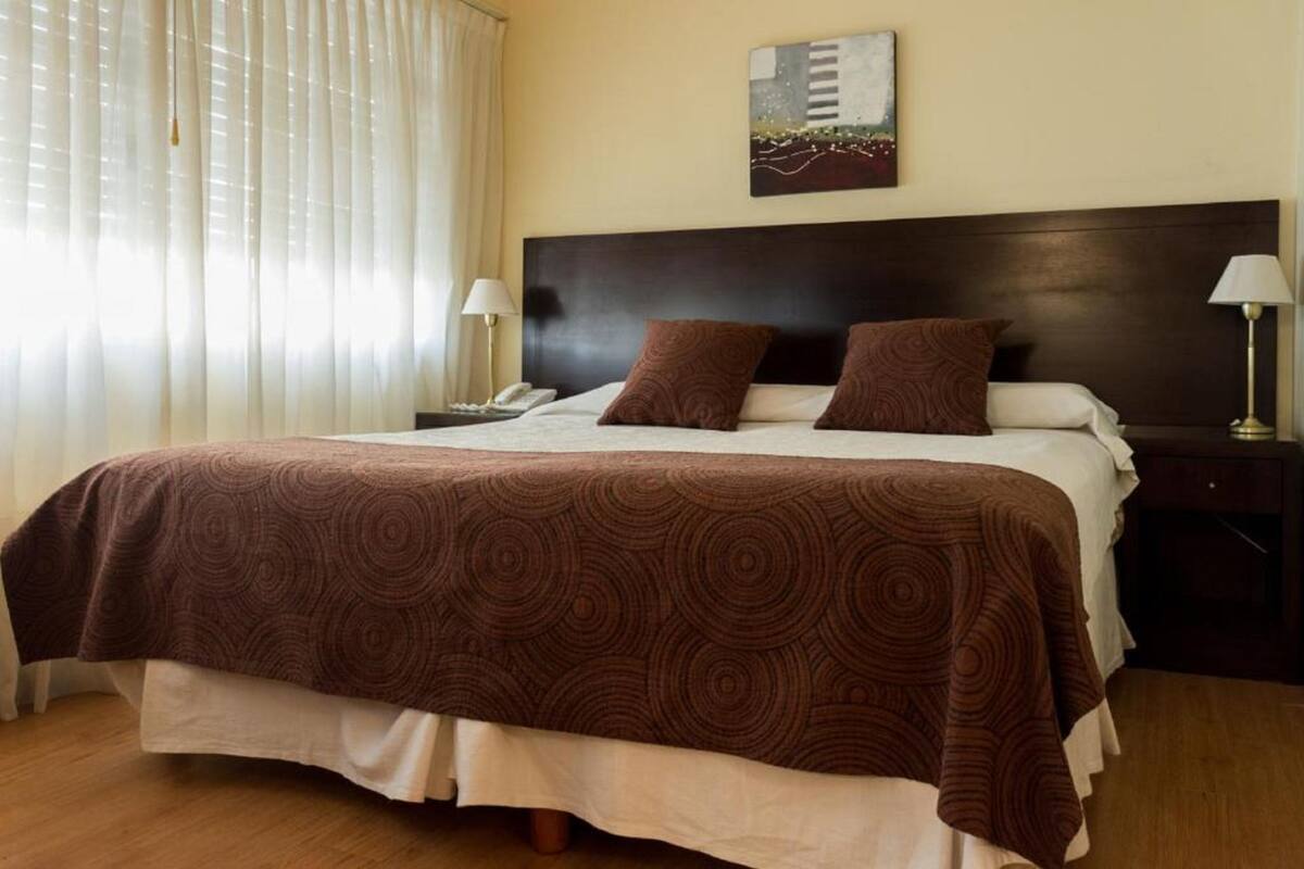 Quarto de hotel com uma cama grande, duas luminárias de cabeceira, um criado-mudo e um quadro na parede.