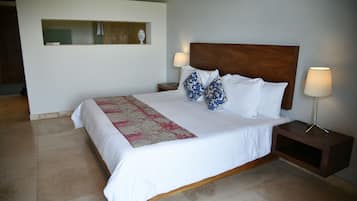 Cama bem arrumada com roupa de cama branca e almofadas azuis estampadas, mesa de cabeceira com abajur e uma pequena estante com um vaso.
