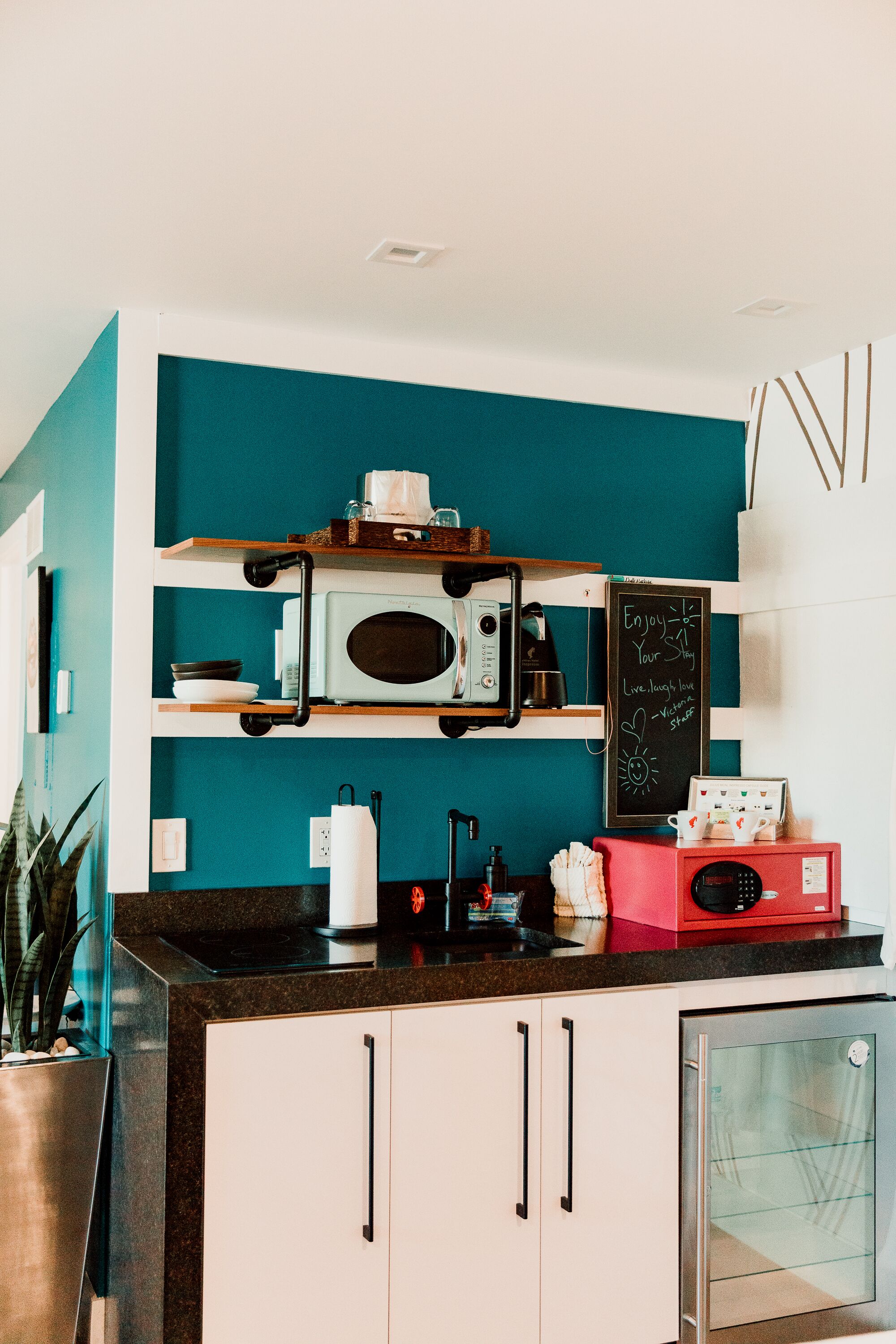 Cozinha com paredes verde-água, micro-ondas, cafeteira vermelha e quadro negro com uma mensagem.