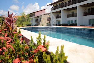 Piscina com borda azulejada, cercada por vegetação e um edifício com varandas.
