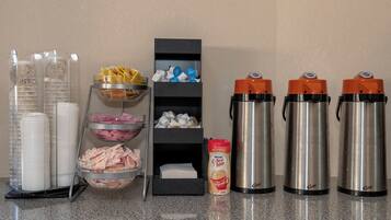 Disposição de buffet com recipientes de vidro transparente para alimentos, estação de café com copos térmicos da marca e pilhas de pratos brancos.