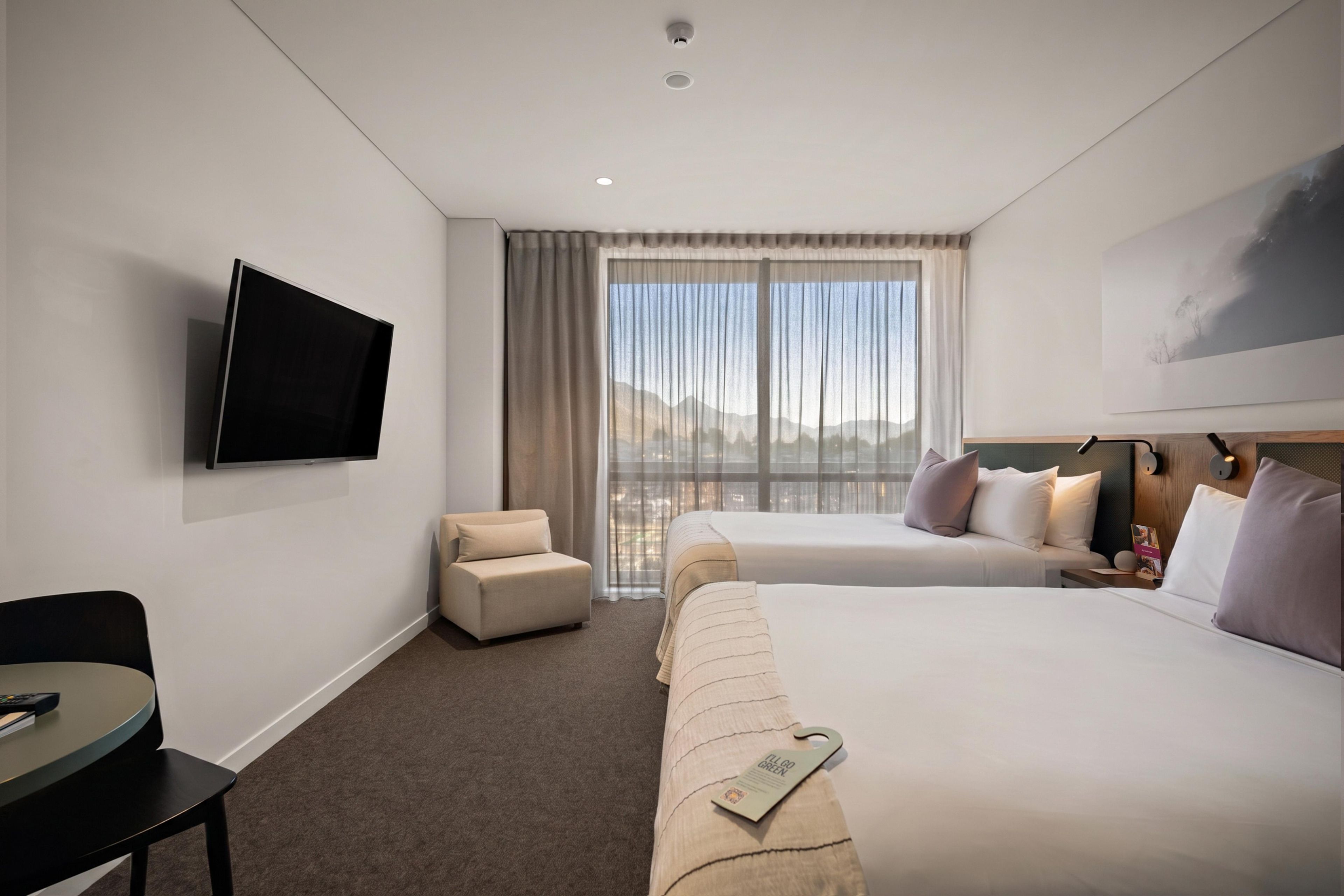 Quarto de hotel com duas camas, televisão, uma cadeira e vista para o exterior.