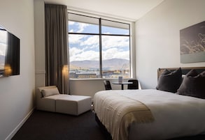 Quarto de hotel com cama, sofá, mesa e cadeira. Janela com vista para montanhas e paisagem urbana.