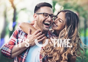 Um casal se abraçando em um quarto com uma janela grande, uma cama e uma mesa de cabeceira.