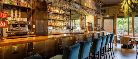 Uma área de bar com balcão, prateleiras cheias de garrafas, copos e uma máquina de café. Há banquetas de bar e uma vista para o exterior através de grandes janelas.