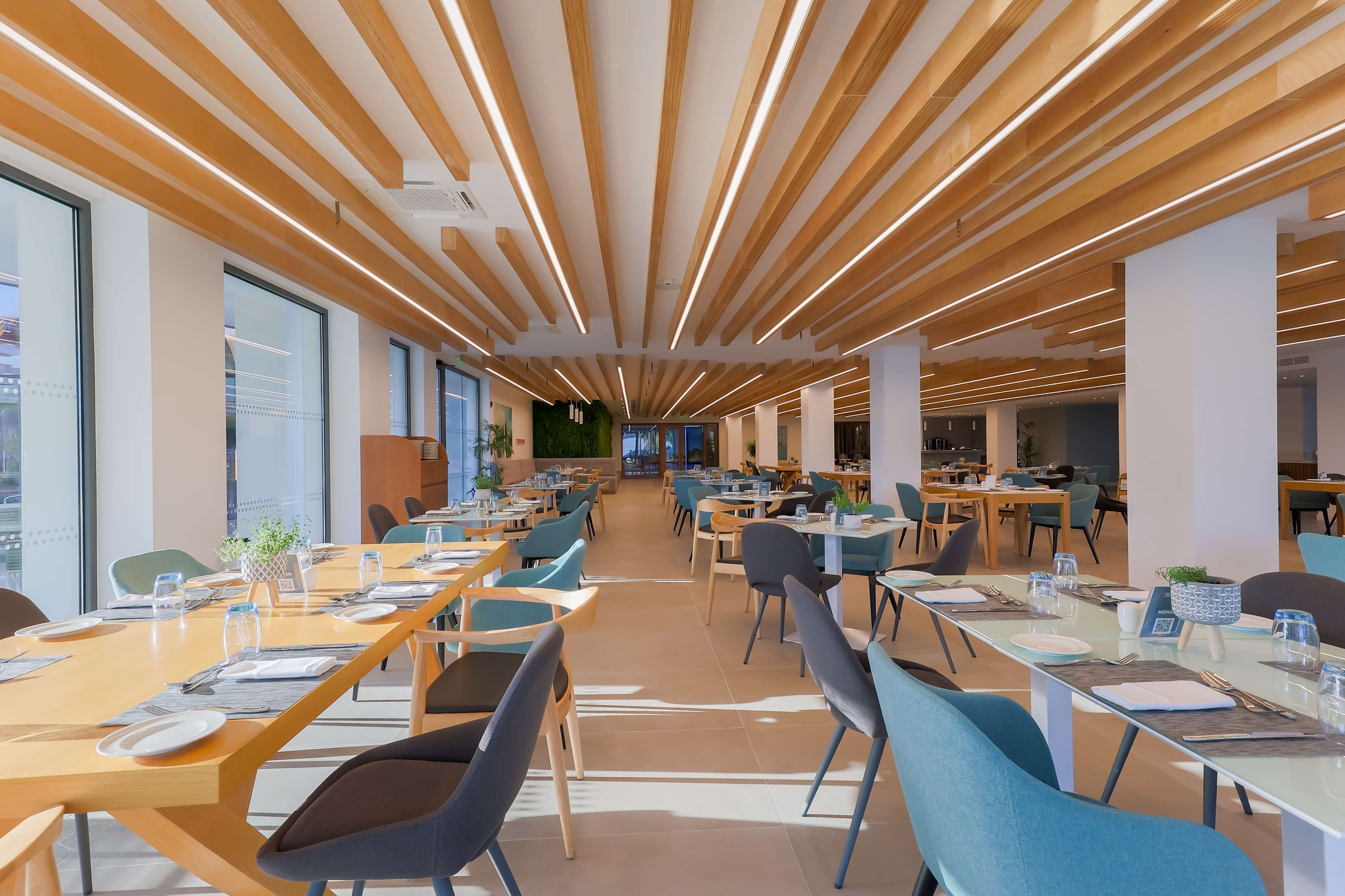 Uma ampla sala de jantar com longas mesas de madeira e cadeiras azuis, grandes janelas e um design moderno no teto.
