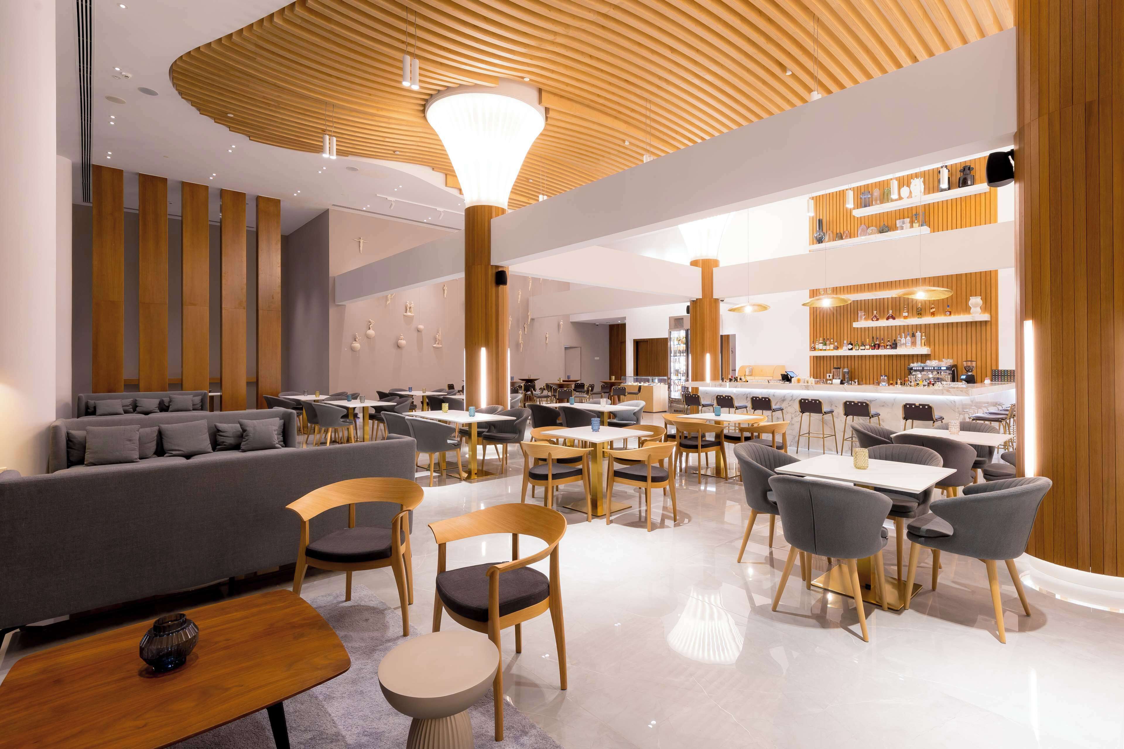 Um saguão de hotel moderno com uma área de estar central, um bar e uma cozinha visível ao fundo.