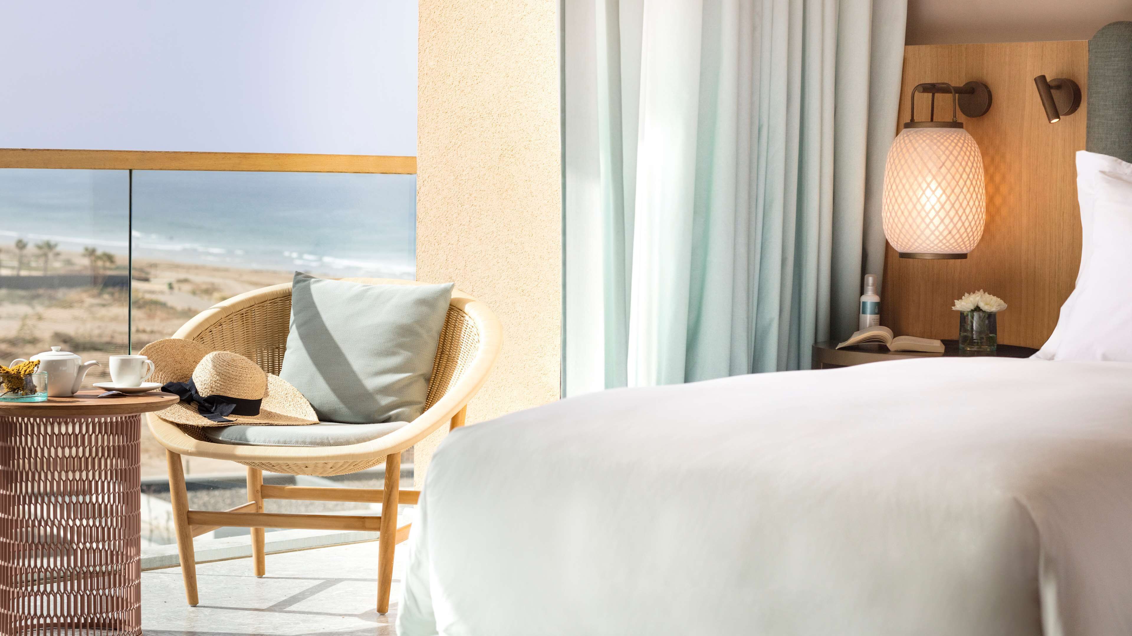 Quarto de hotel com cama, cadeira de vime com almofadas, mesa de cabeceira com abajur e vista para a praia.