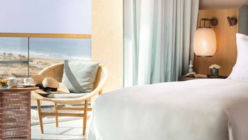 Quarto de hotel com cama, cadeira de vime com almofadas, mesa de cabeceira com abajur e vista para a praia.