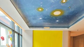 Espaço infantil com piscina de bolinhas, escorregadores e um mural no teto representando o sistema solar.