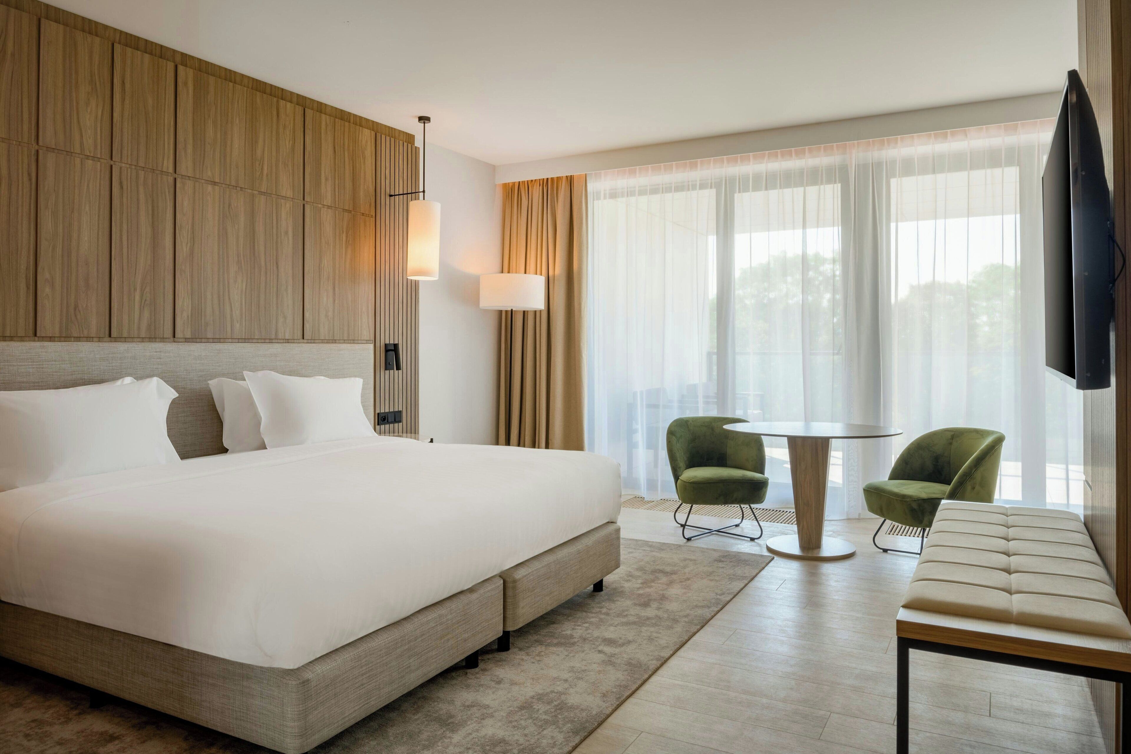 Quarto de hotel moderno com uma cama grande, televisão, área de estar com duas cadeiras e uma janela ampla com cortinas.