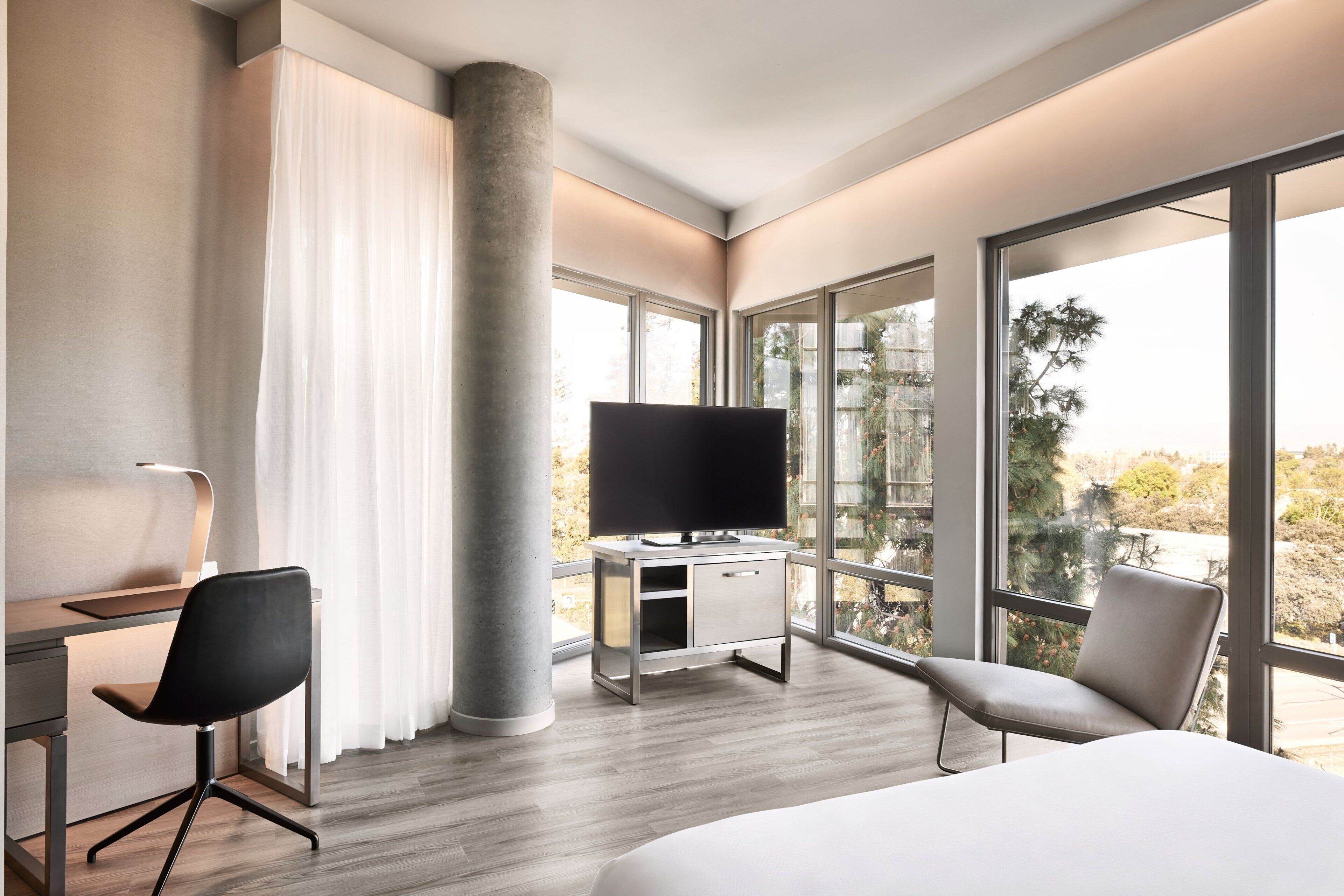 Quarto de hotel moderno com uma cama grande, uma escrivaninha com cadeira, uma televisão sobre um suporte e grandes janelas com vista para o exterior.