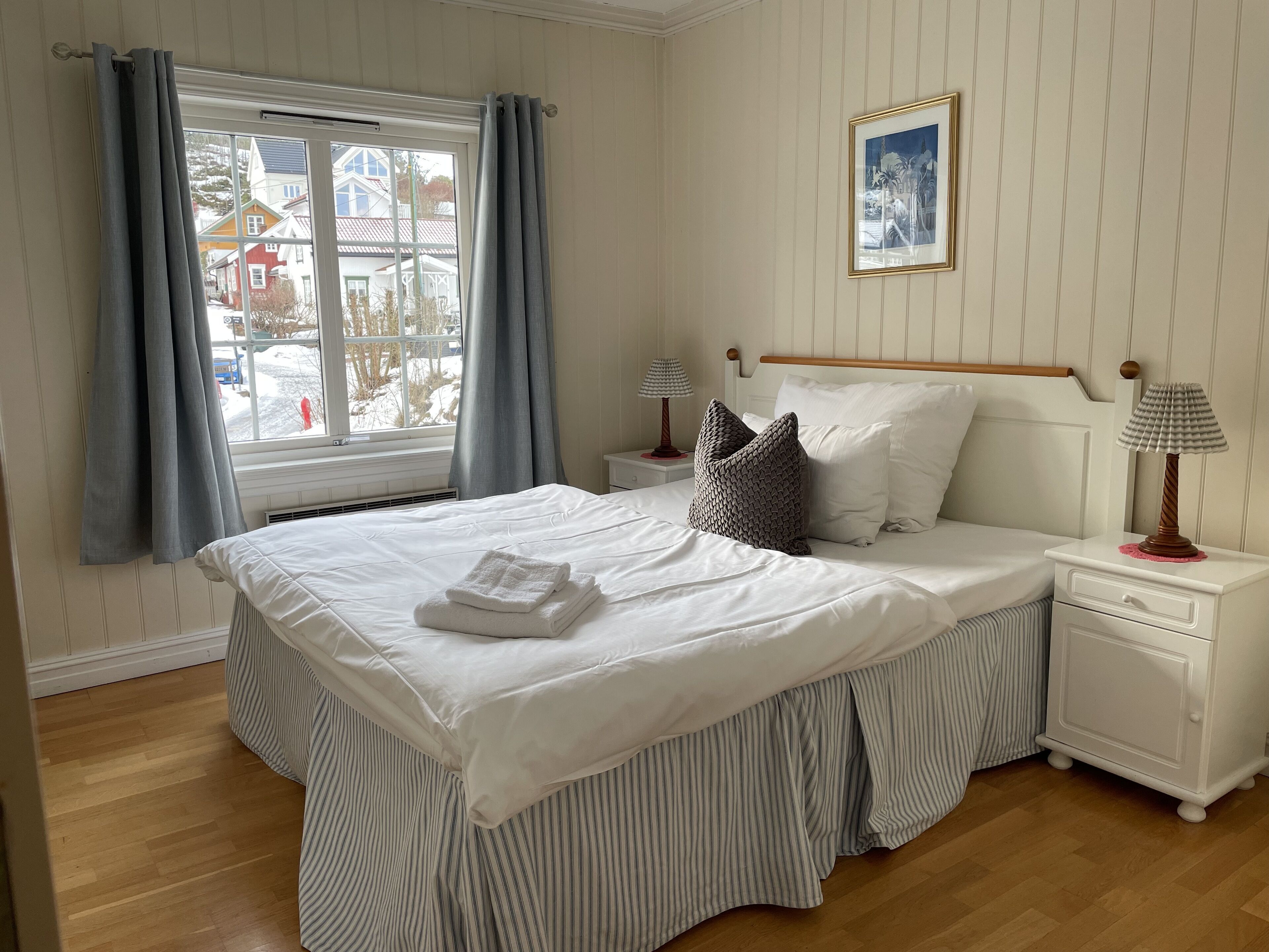 Uma cama bem arrumada com roupa de cama branca e cortinas listradas, um abajur e um quadro na parede.