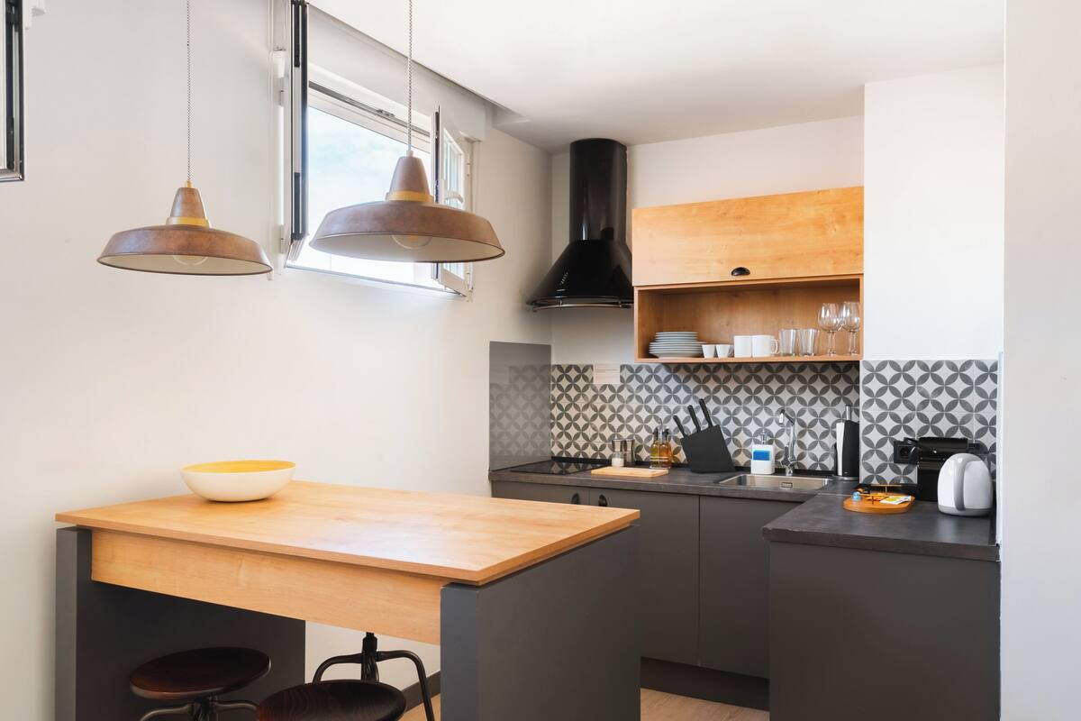 Uma cozinha compacta com uma mesa de madeira, dois banquetas altas, luminárias pendentes, um exaustor preto e um revestimento de parede estampado.