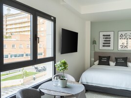 Quarto de hotel com uma janela grande, uma cama, uma TV e uma pequena mesa com uma planta.