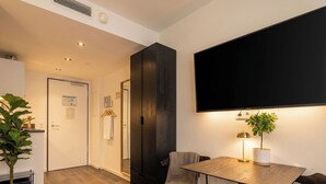 Quarto moderno com mesa de madeira, cadeiras, planta em vaso, armário preto e TV na parede.
