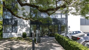 Entrada de um edifício moderno com porta de vidro, placa de identificação e área de estacionamento com carros.