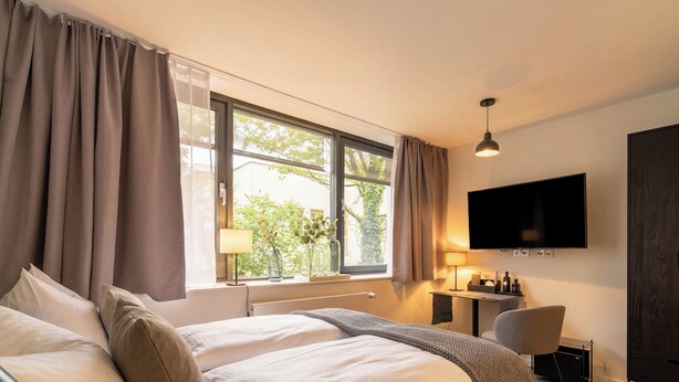 Quarto de hotel com cama, escrivaninha, cadeira, televisão e janela com cortinas.