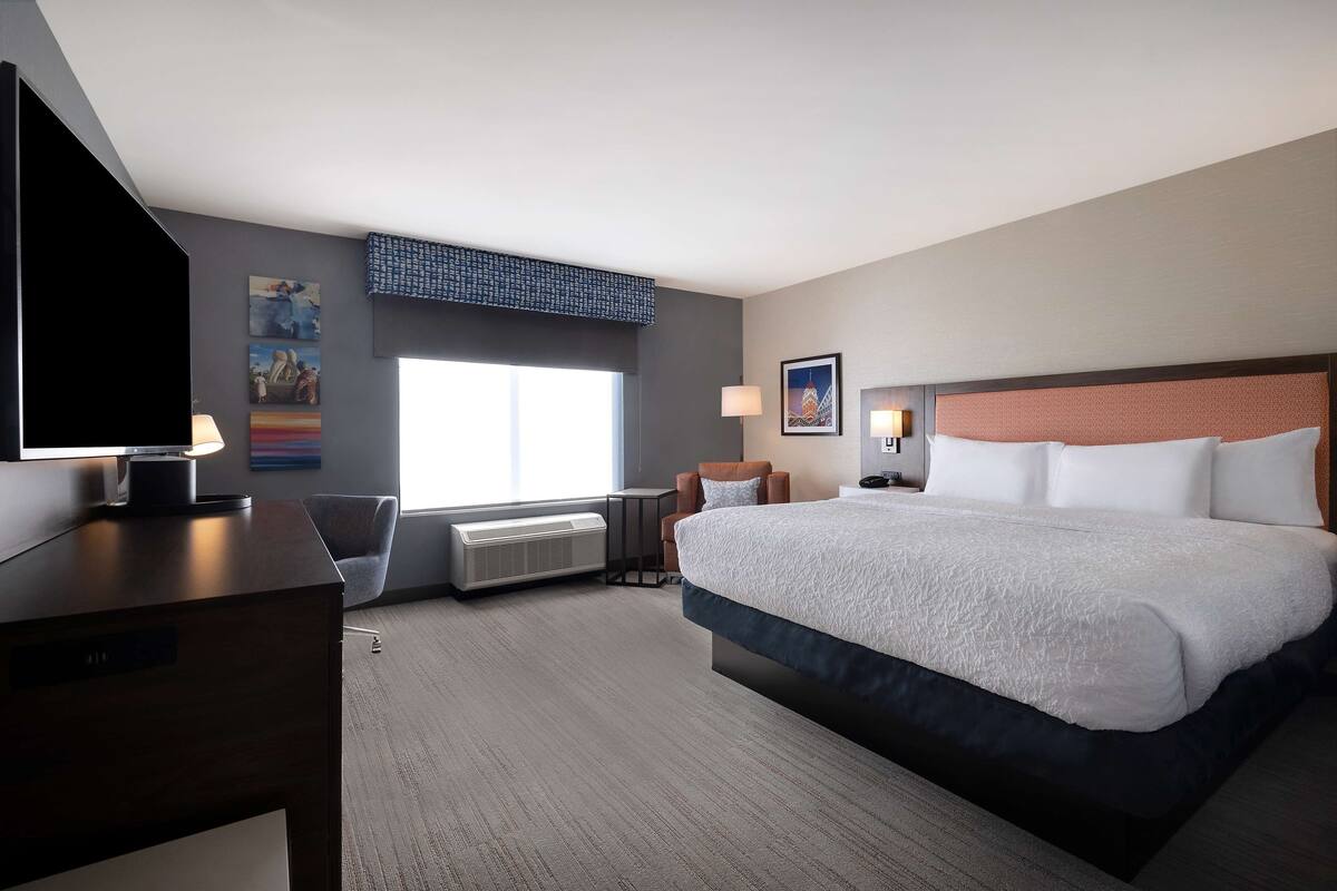 Quarto de hotel com uma cama grande, escrivaninha com televisão de tela plana, cadeira, janela com persianas e ar-condicionado fixado na parede.