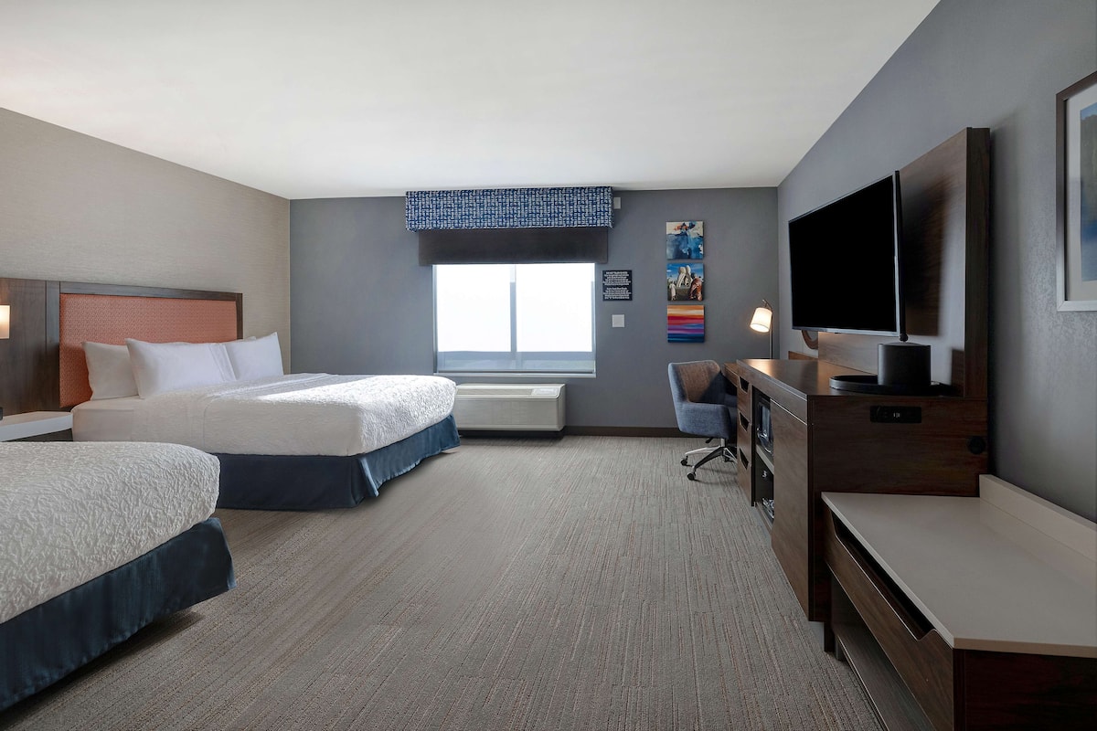 Quarto de hotel com duas camas, uma escrivaninha, televisão e uma janela grande.