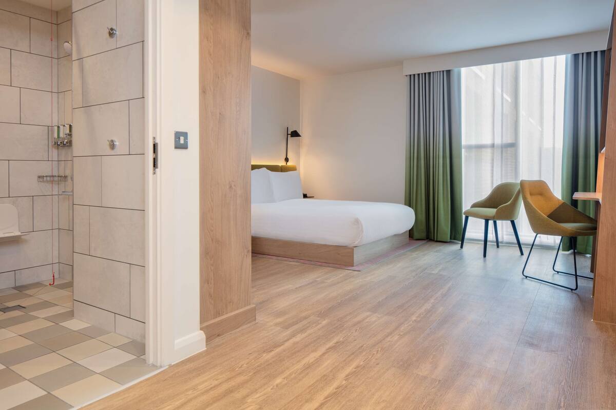 Quarto de hotel com cama, escrivaninha, duas cadeiras, área de chuveiro e uma janela grande com cortinas.