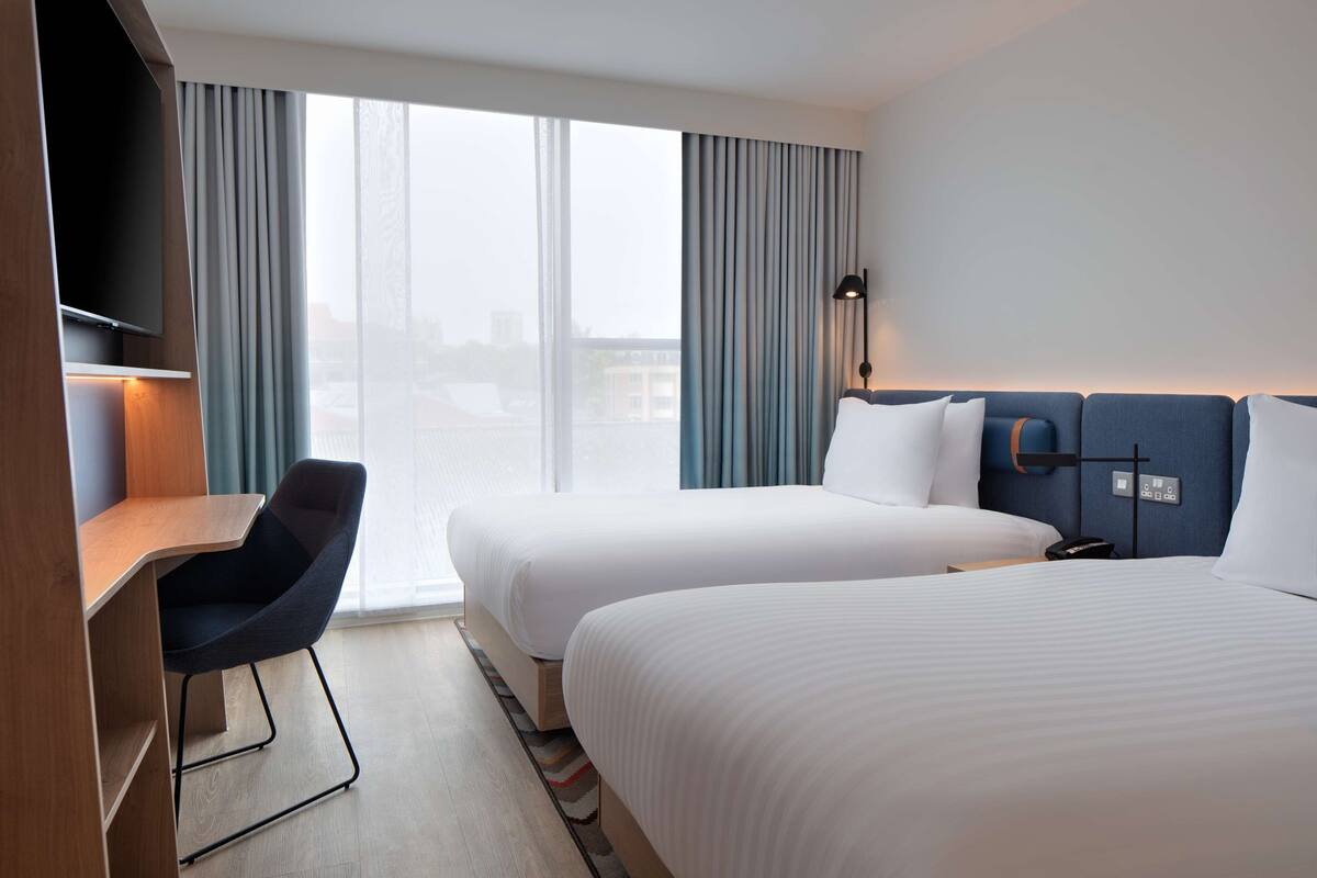 Quarto de hotel com duas camas, uma escrivaninha, uma cadeira, televisão e uma janela grande com cortinas.