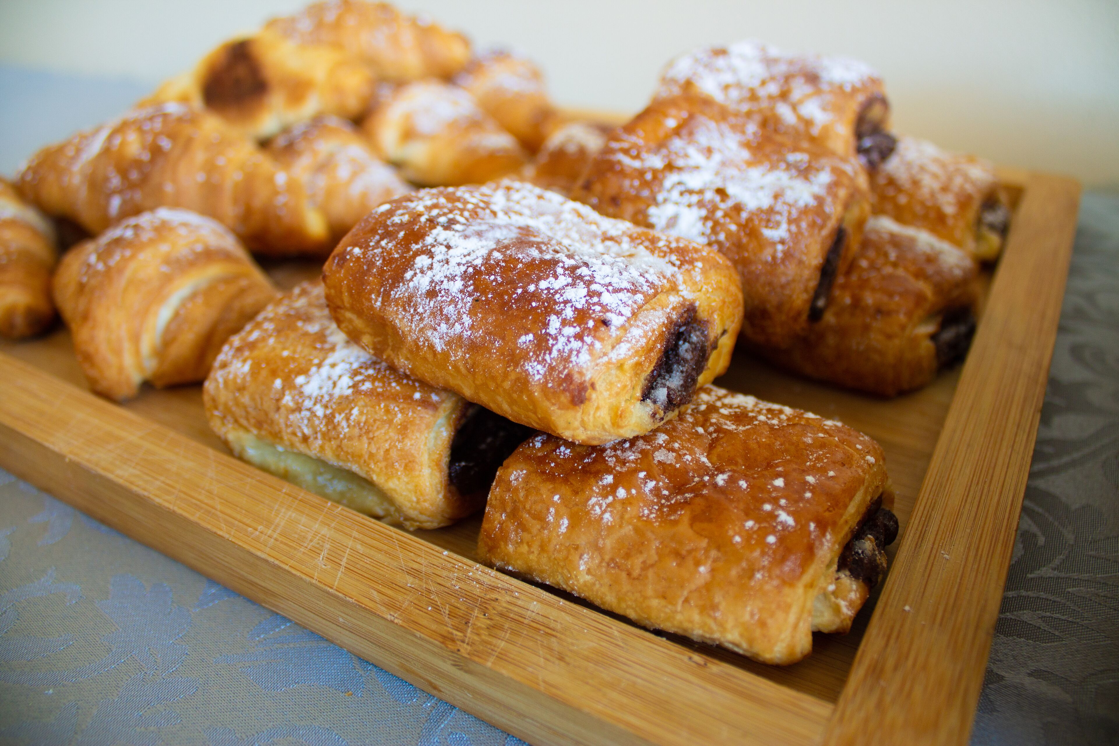 Uma bandeja de madeira com diversos doces, incluindo croissants e pasteis recheados com chocolate, polvilhada com açúcar em pó.