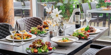 Mesa posta com pratos de comida, copos e uma garrafa de vinho, cercada por cadeiras e vista para o verde.