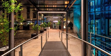 Corredor moderno de hotel com paredes de vidro, grades de metal e plantas em vasos.