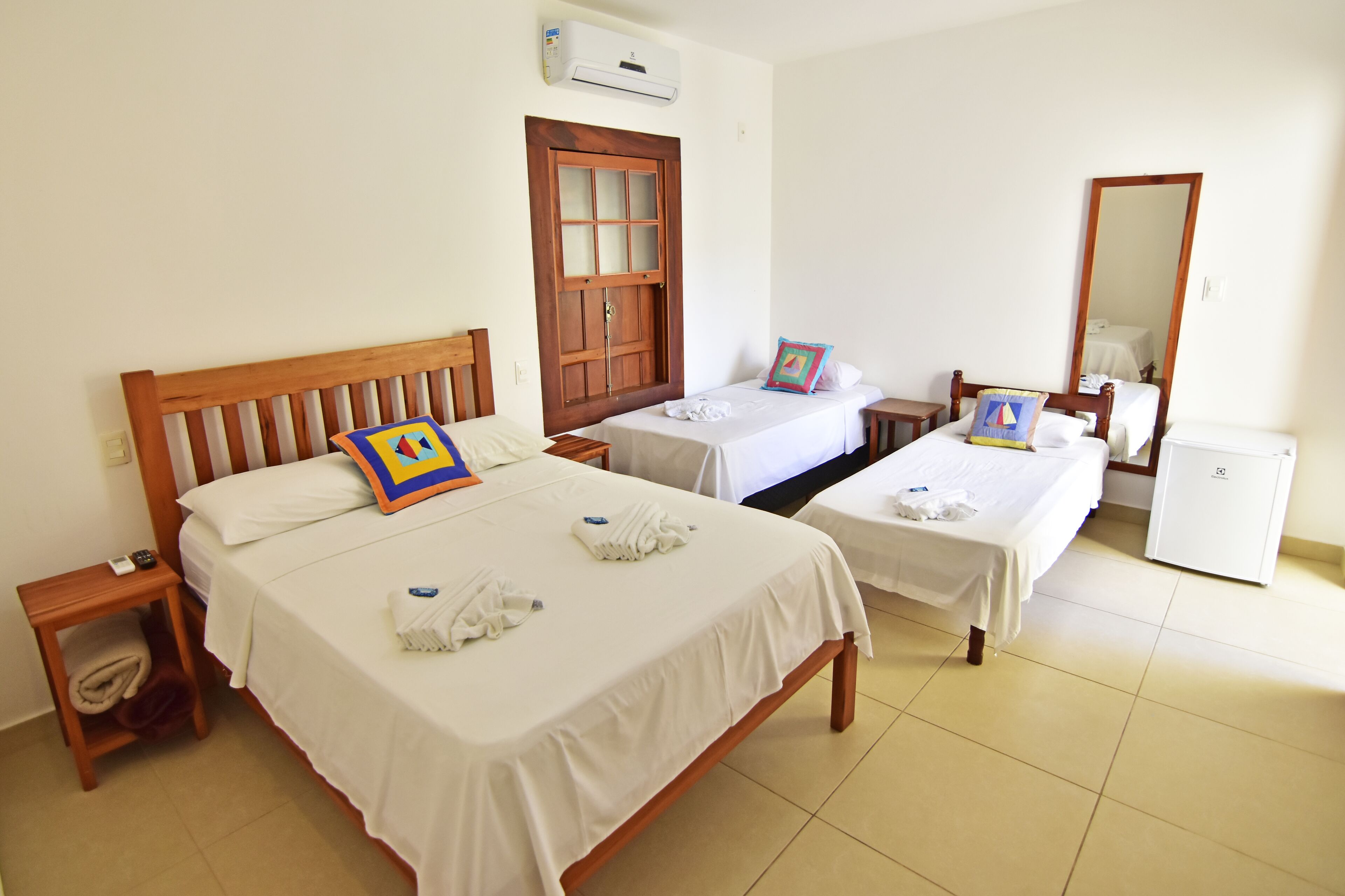 Quarto de hotel com duas camas de solteiro, cabeceira de madeira, mesa de cabeceira pequena com telefone, criado-mudo, espelho e geladeira branca.