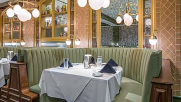 Restaurante com assentos estofados em verde, toalhas brancas e luminárias pendentes esféricas.