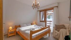 Quarto com cama de madeira, mesa de cabeceira com abajur, cadeira com manta e vista para um edifício lá fora.