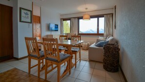 Uma área de refeições com cadeiras de madeira e uma mesa, um sofá com almofadas, uma televisão na parede e vista para montanhas pelas janelas.
