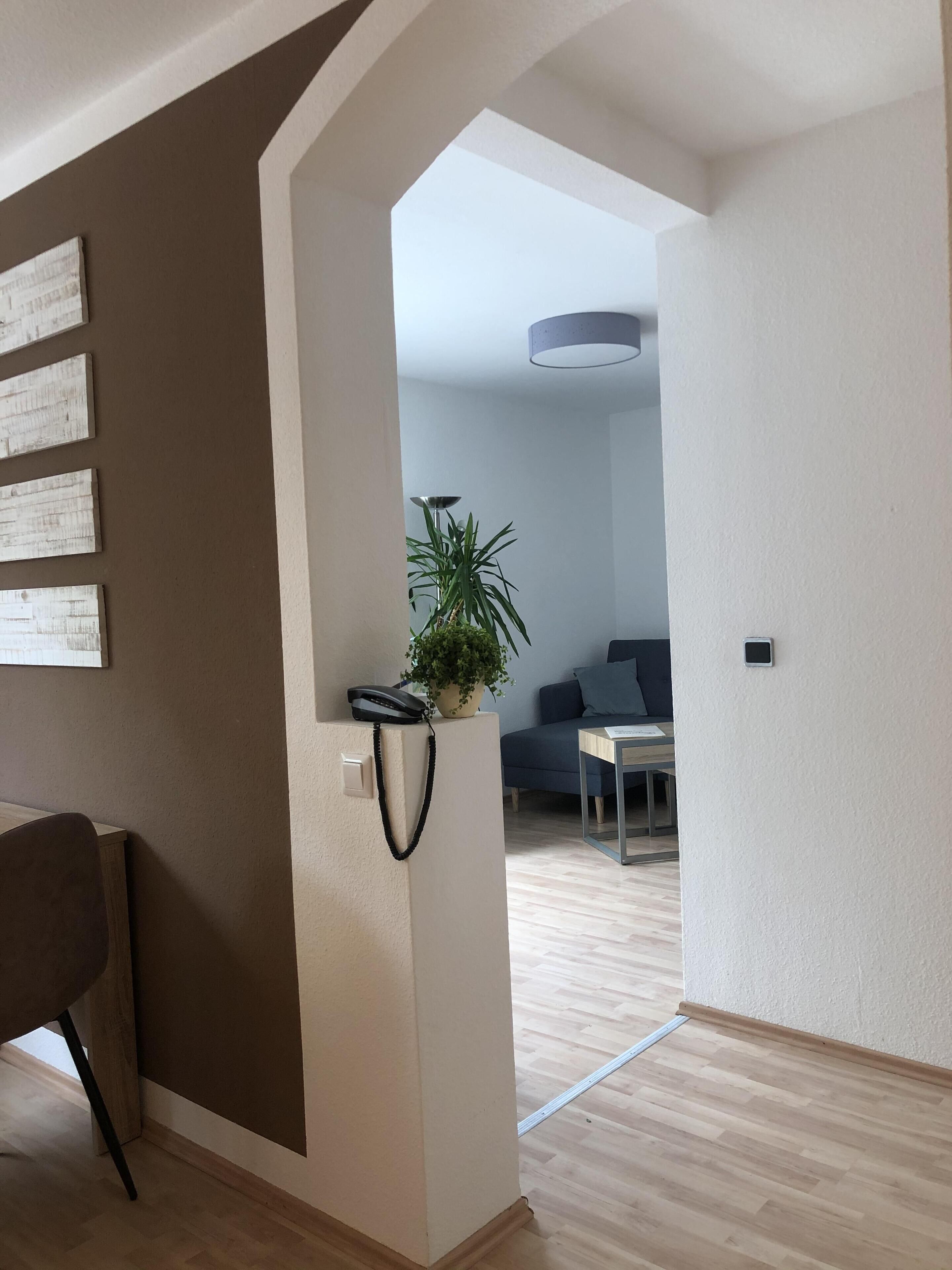 Uma sala de estar moderna com uma parede em tom marrom escuro, piso de madeira, sofá azul, uma mesinha e um vaso com planta.