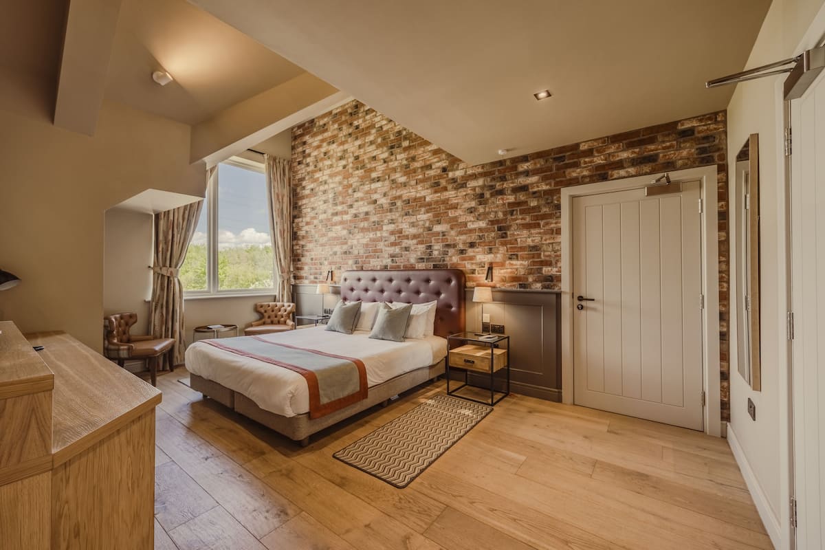 Um quarto com uma cama grande, parede de tijolos, piso de madeira, mesa de cabeceira, luminária e porta.