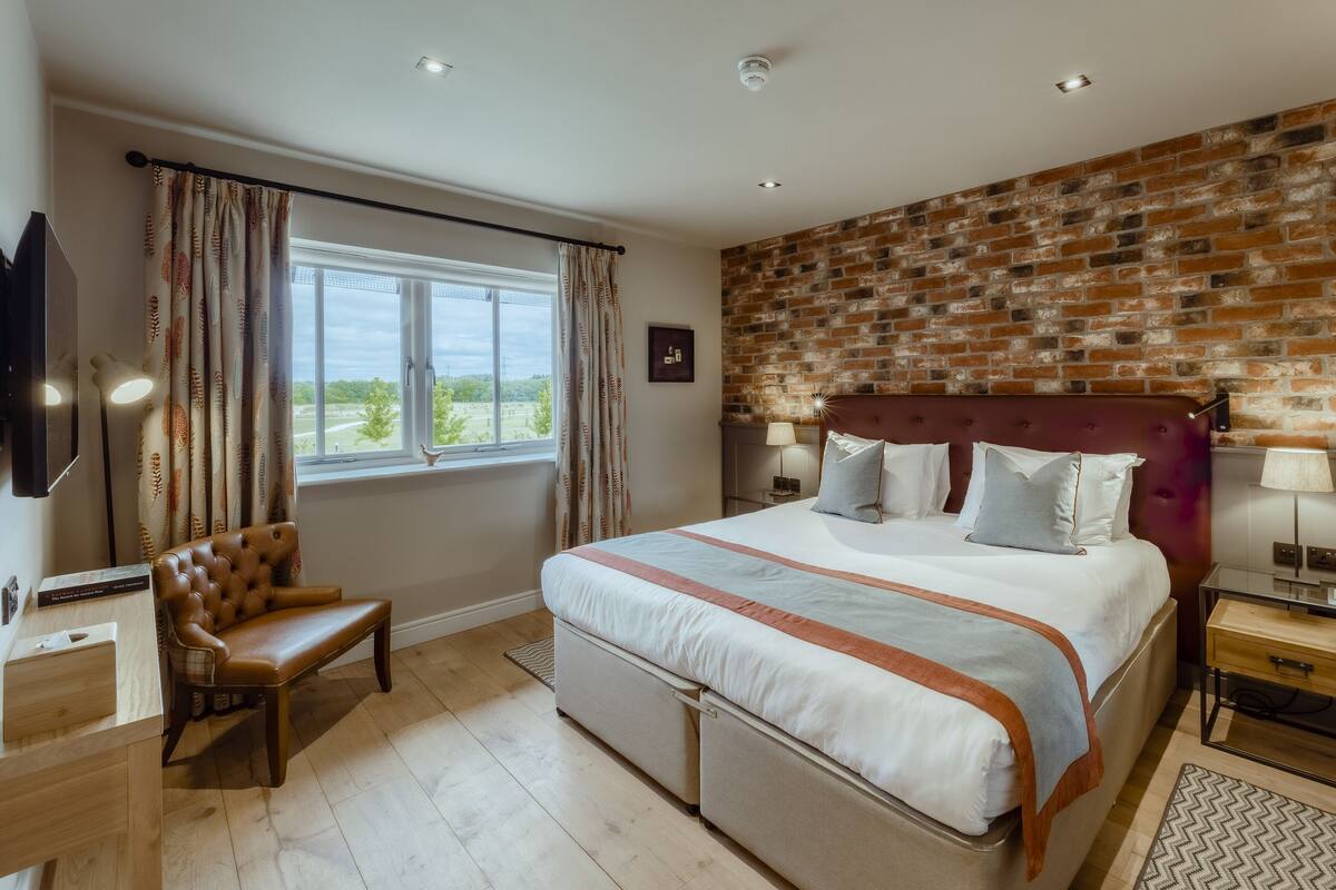 Quarto de hotel com uma cama grande, uma parede de tijolos, uma escrivaninha com cadeira e uma janela com cortinas.