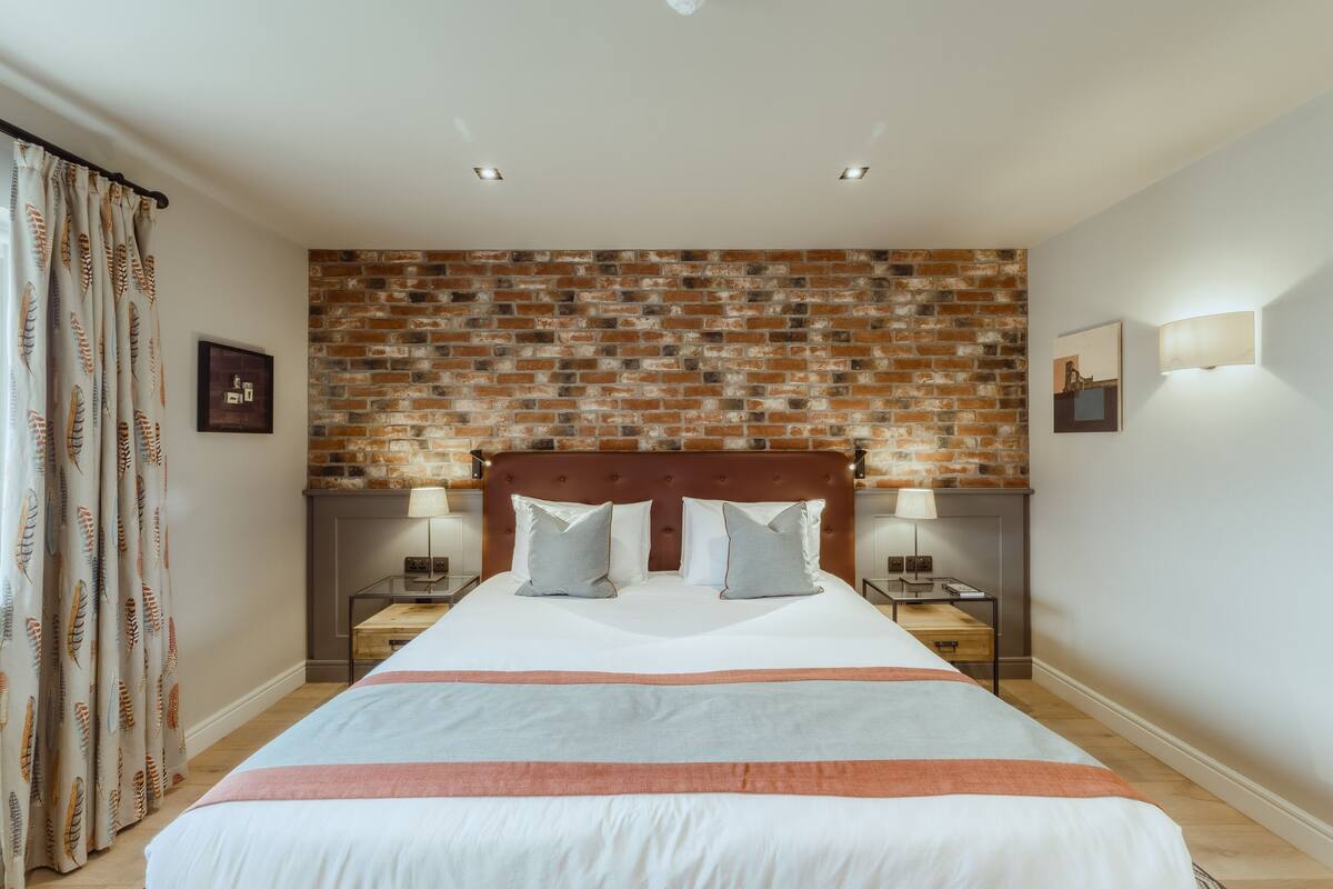 Quarto de hotel com parede em padrão de tijolos, uma cama grande com cabeceira de madeira, duas mesas de cabeceira com abajures e cortina com padrão de folhas.