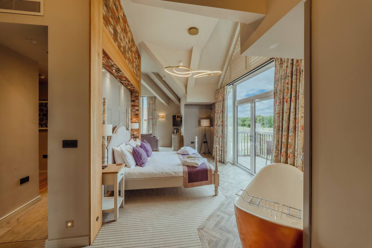 Quarto de hotel moderno com uma cama grande, um espaço de estar com uma poltrona, vista para o exterior e uma varanda.