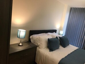 Quarto de hotel com cama, duas luminárias de cabeceira, mesa de cabeceira e janela com persianas.