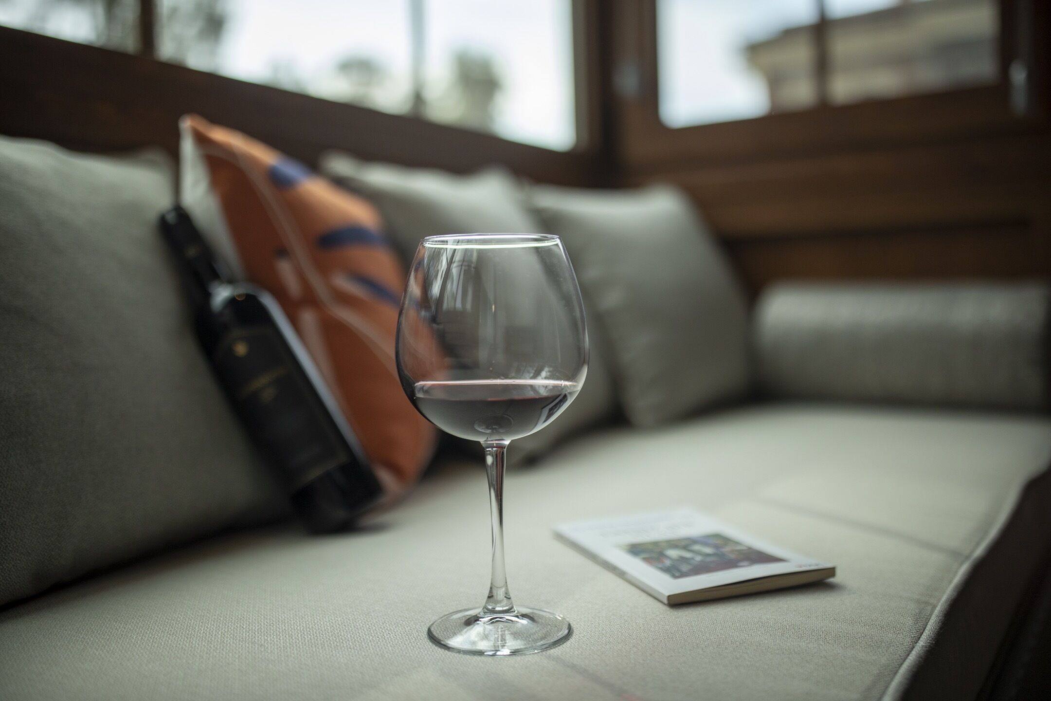 Um copo de vidro com vinho tinto em um sofá acolchoado, com um livro e uma bolsa ao fundo.